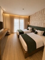 Quarto de hotel com duas camas, televisão de tela plana, mesa de cabeceira e uma janela grande com cortinas.