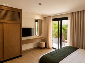 Quarto de hotel com uma cama, televisão, espelho, um pequeno banquinho e uma porta de vidro deslizante que dá para uma varanda com plantas.