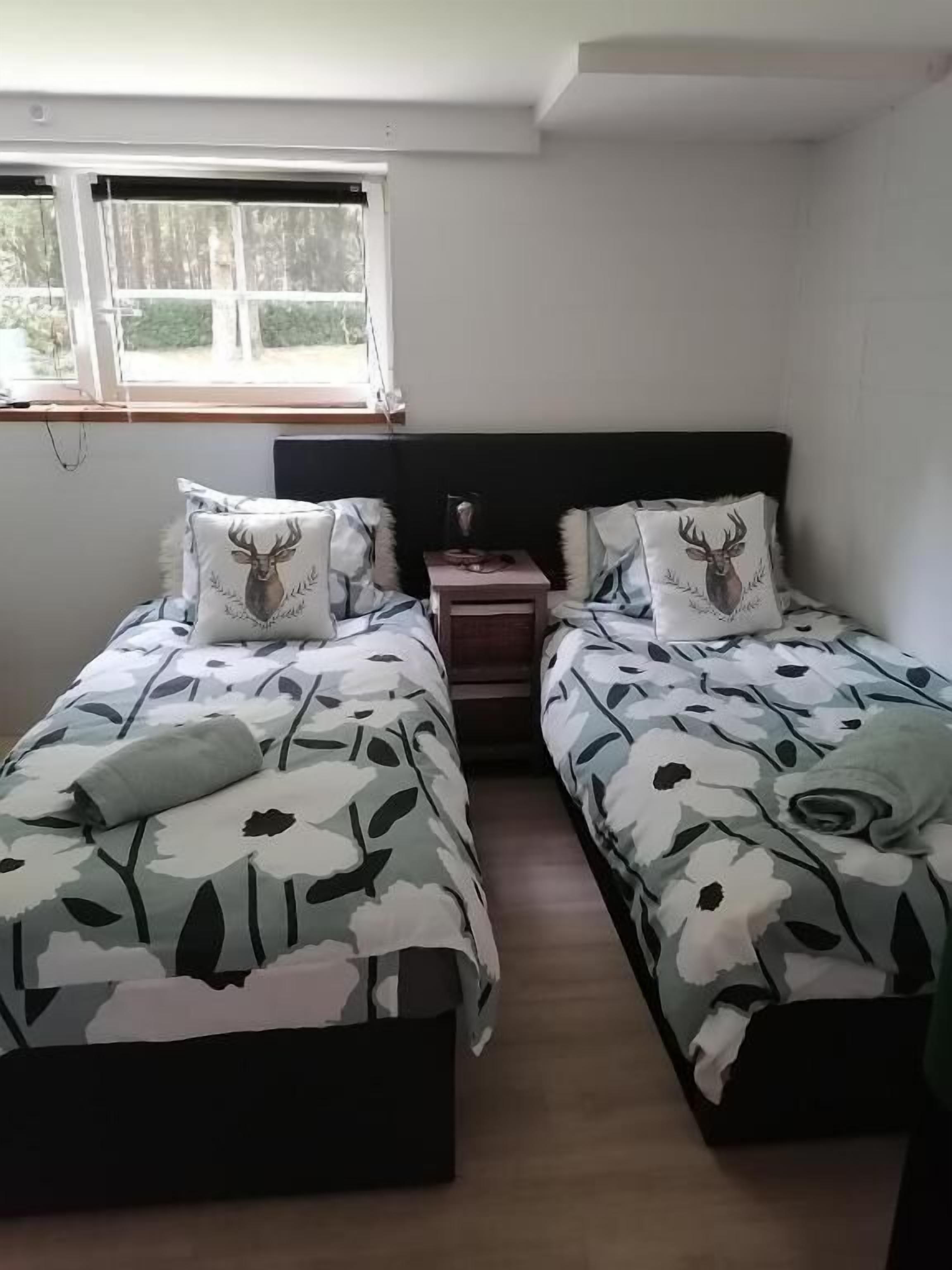 Quarto com duas camas, cada uma com roupa de cama estampada com flores e almofadas com tema de veados. Há uma pequena mesa de cabeceira com uma luminária entre as camas. Uma janela com vista para árvores é visível.