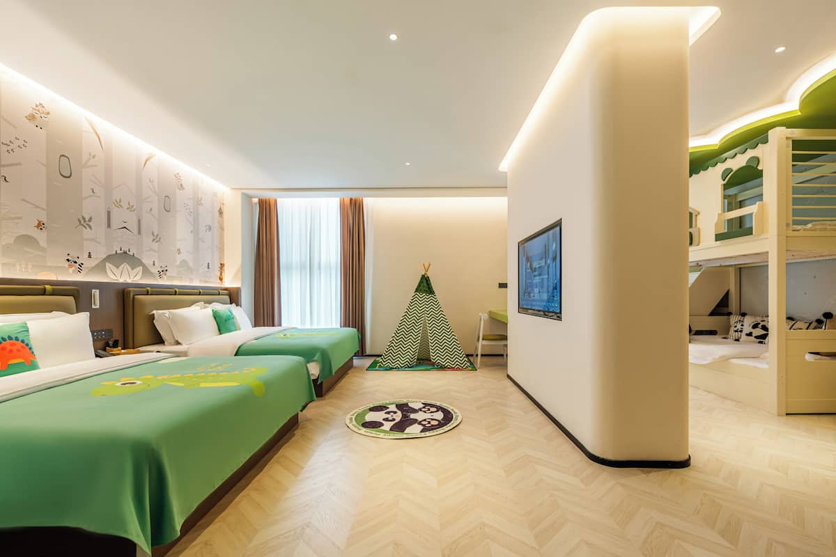 Quarto de hotel com duas camas, parede com detalhes verdes, uma pequena árvore decorativa, um tapete redondo e uma cama beliche no canto.