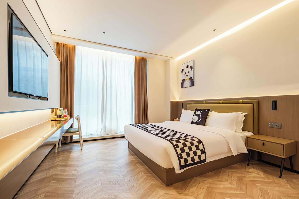 Quarto de hotel com cama, mesa de cabeceira, televisão e uma grande janela com cortinas.