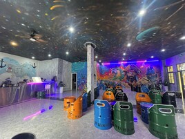 Quarto com um mural de tema espacial, um balcão de recepção e barris coloridos com a inscrição 'WIN HOUSE'.