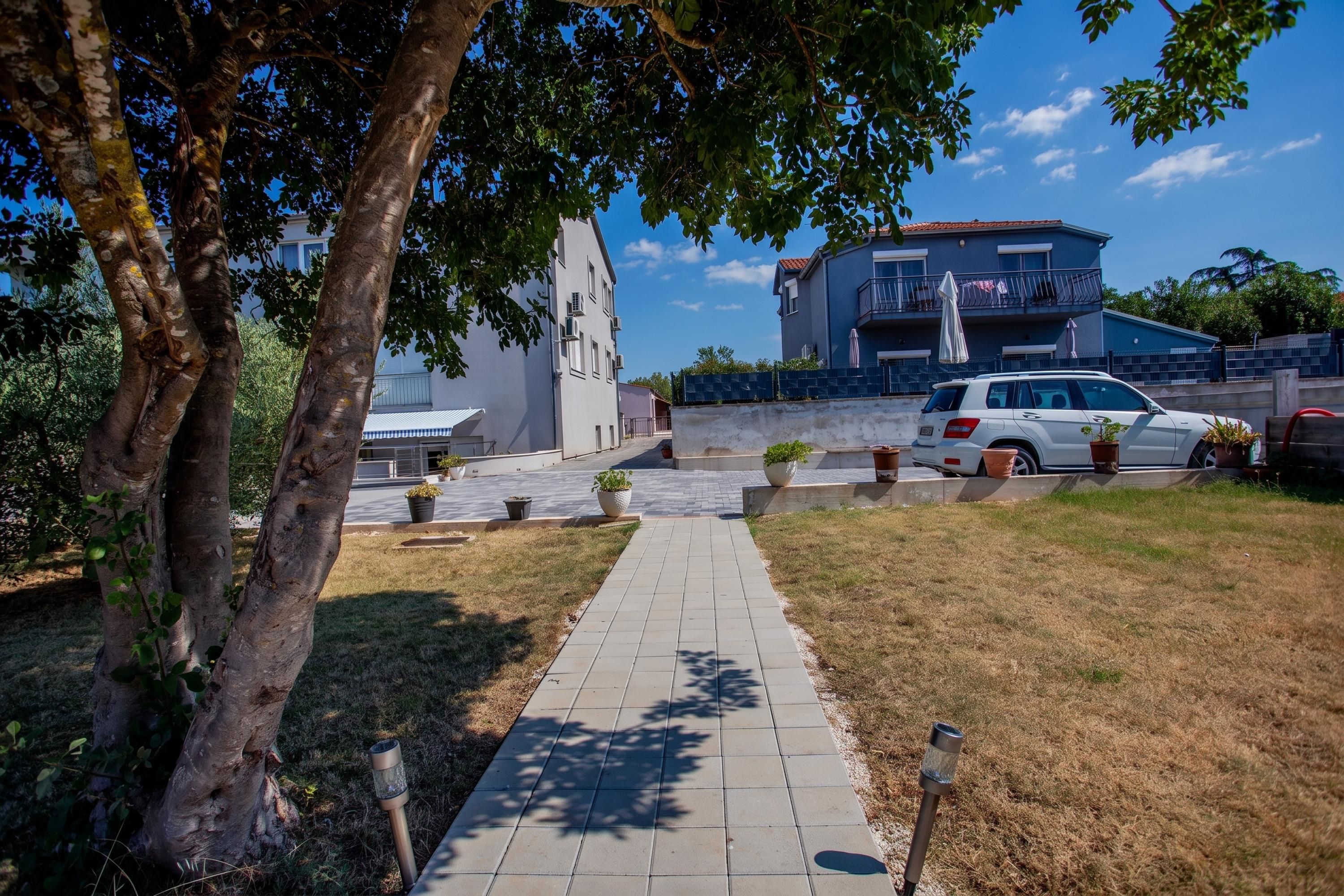 Uma trilha pavimentada leva a um edifício com varanda, rodeado por árvores e plantas em vasos.