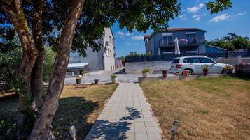 Uma trilha pavimentada leva a um edifício com varanda, rodeado por árvores e plantas em vasos.