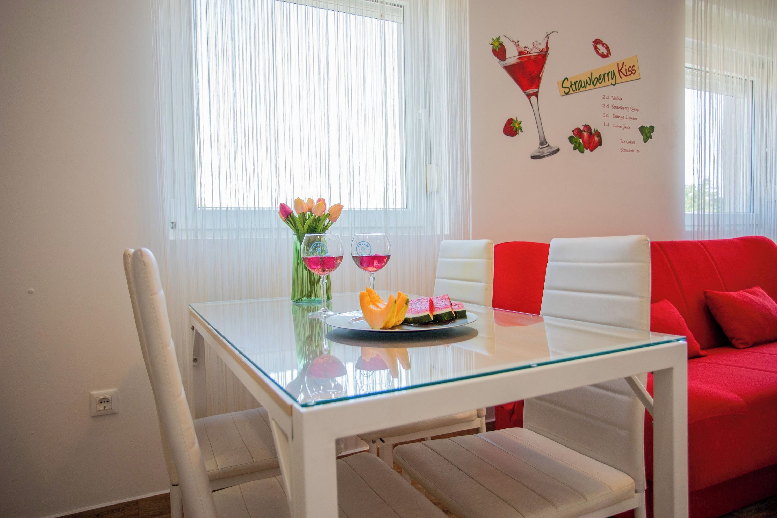 Uma sala de jantar com mesa de vidro, cadeiras brancas, um sofá vermelho e um objeto decorativo na parede.