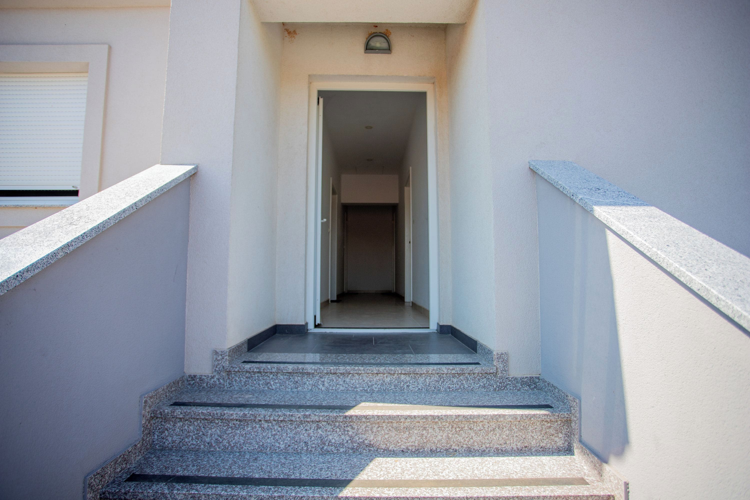 Degraus de concreto levam a uma porta, com corrimão de um lado e uma janela com persianas do outro.