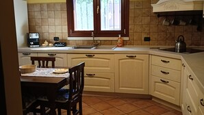 Cozinha com mesa de jantar, cadeiras, máquina de café, chaleira e janela com cortinas.