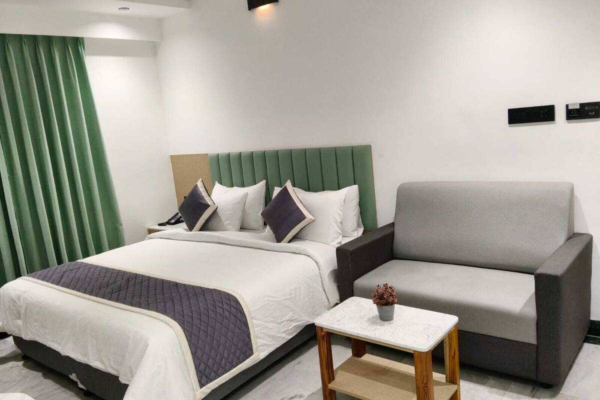 Quarto de hotel moderno com uma cama, uma poltrona cinzenta, uma mesinha e uma cortina verde.
