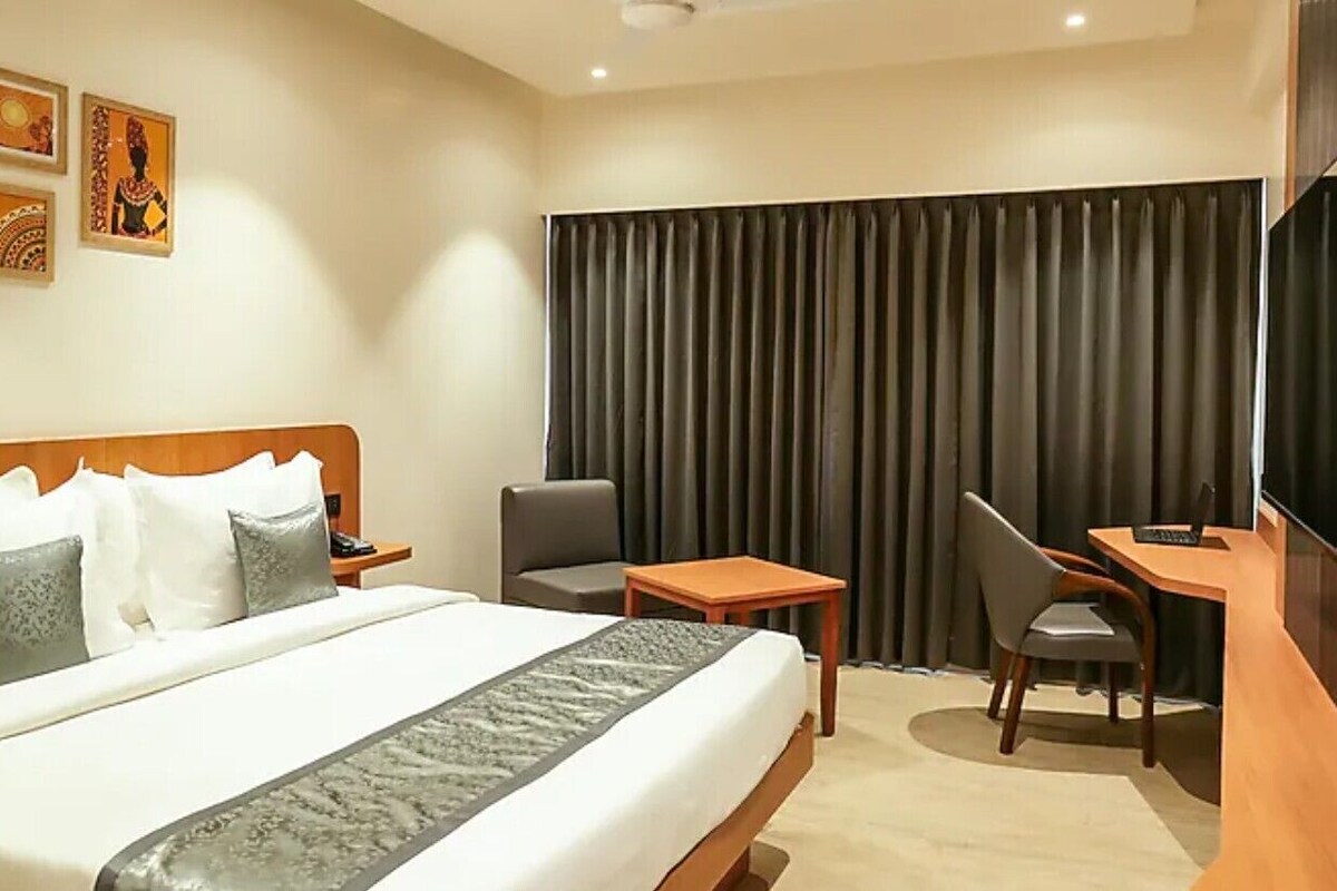 Quarto de hotel com cama, escrivaninha, cadeira, televisão e uma grande janela com cortinas.