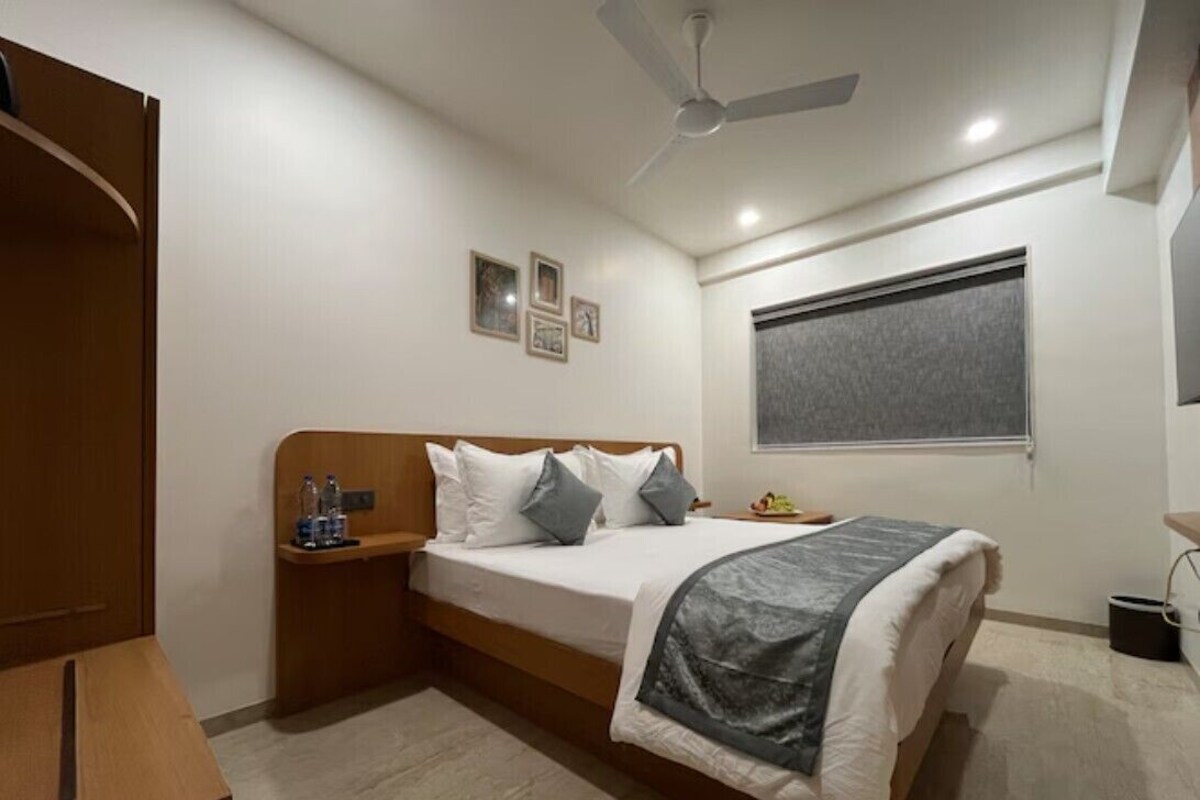 Quarto de hotel com cama, mesa de cabeceira, prateleira na parede, ventilador de teto, janela e um pequeno cesto de lixo.
