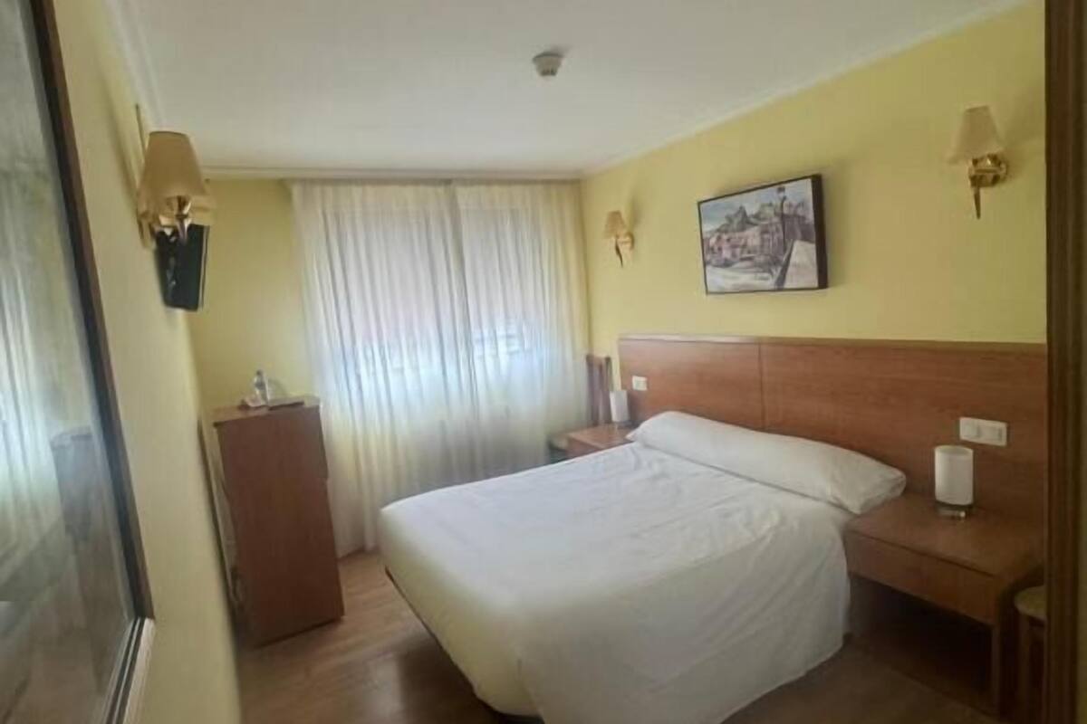 Quarto de hotel com cama, mesa de cabeceira, luminária, quadro na parede e janela com cortinas.