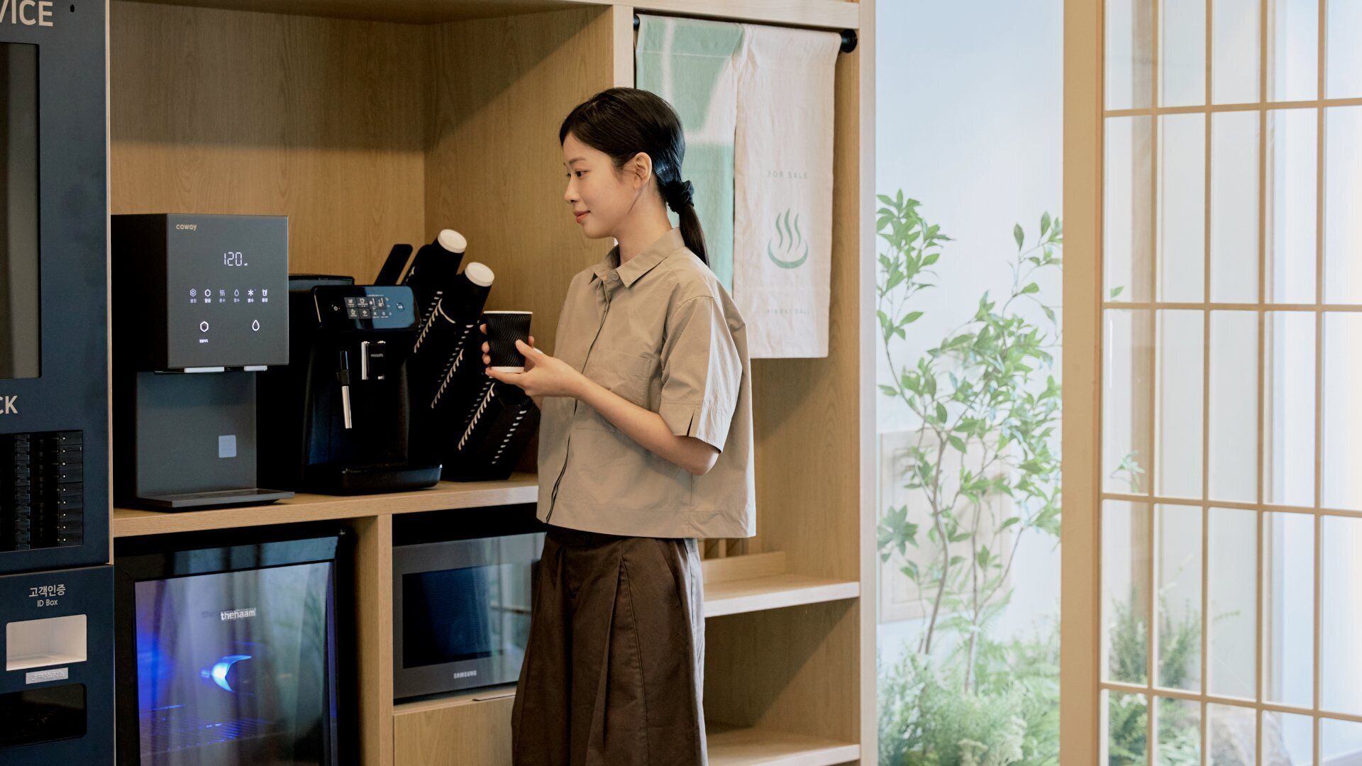 Uma pessoa em frente a uma máquina de café e uma geladeira Samsung, com uma prateleira de madeira e uma planta ao fundo.