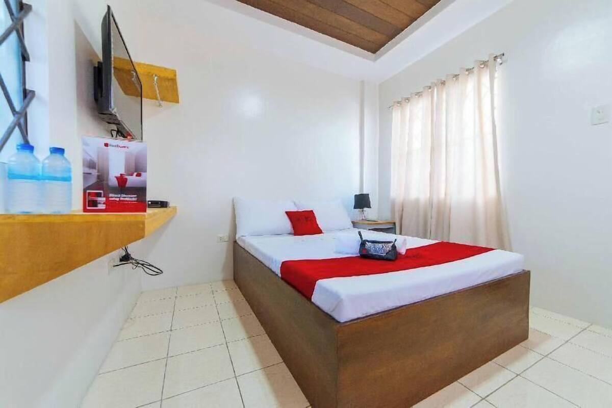 Quarto com cama de madeira, paredes brancas, teto de madeira, TV fixada na parede e janela com cortinas.