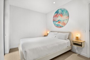 Quarto com cama, luminárias de cabeceira, uma pintura abstrata na parede e mesa de cabeceira.