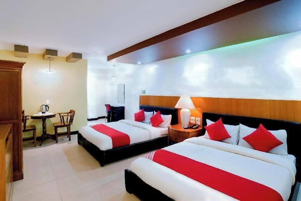 Quarto de hotel com duas camas, uma mesa pequena com cadeiras, uma luminária e uma cabeceira de madeira.