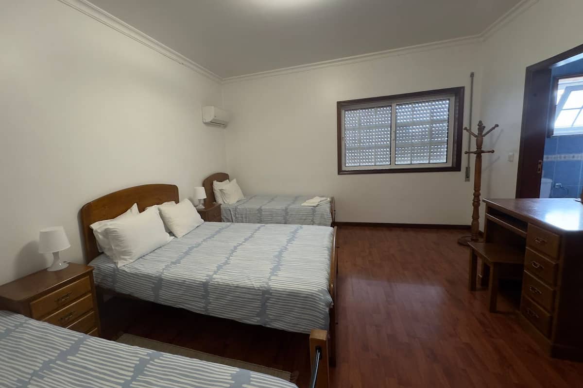 Quarto com duas camas, uma escrivaninha, uma cadeira, janela com persianas e ar-condicionado fixado na parede.