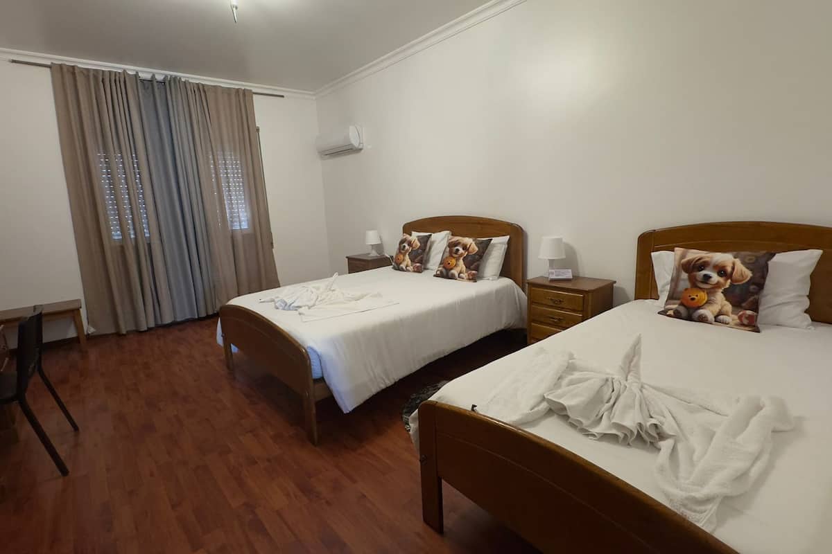 Quarto de hotel com duas camas, uma escrivaninha, uma cadeira, um guarda-roupa e uma janela com cortinas.
