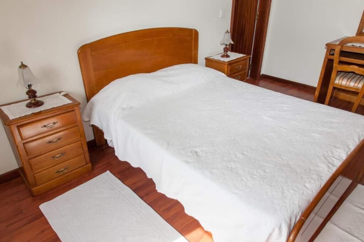 Quarto com cama de madeira, mesa de cabeceira com luminária, cômoda de madeira e uma cadeira.