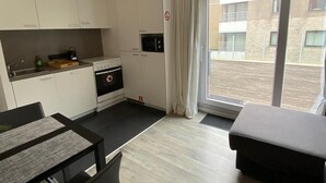 Uma cozinha compacta com armários brancos, micro-ondas e forno pequeno. Há uma mesa de jantar com cadeiras e um banco. O espaço possui uma grande porta de vidro deslizante que dá para uma área externa.