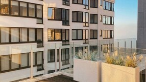 Um edifício moderno de vários andares com grandes janelas, um terraço no telhado com piso de madeira e vasos com plantas.