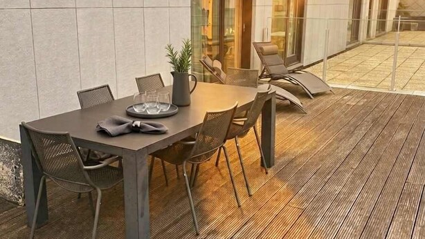 Uma área de refeições ao ar livre com mesa, cadeiras, uma planta em vaso e uma vista de um edifício ao fundo.