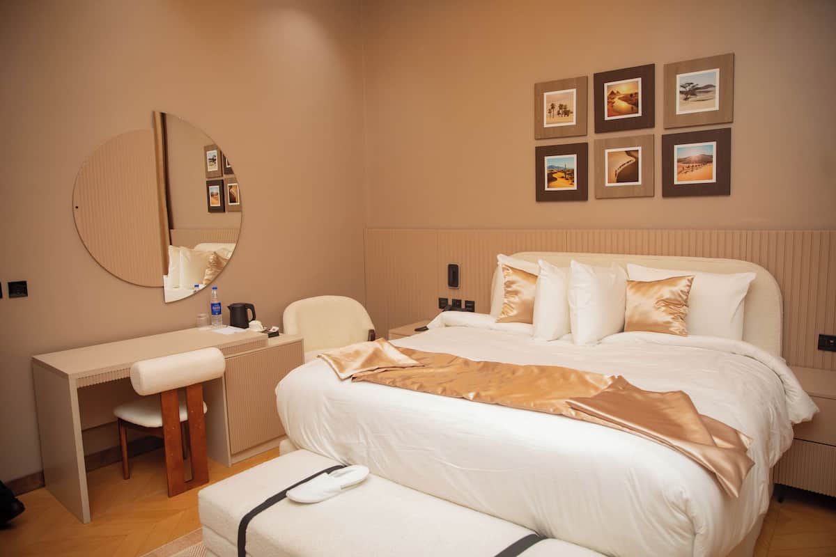 Quarto de hotel com cama, escrivaninha, cadeira, espelho redondo e quadros nas paredes.