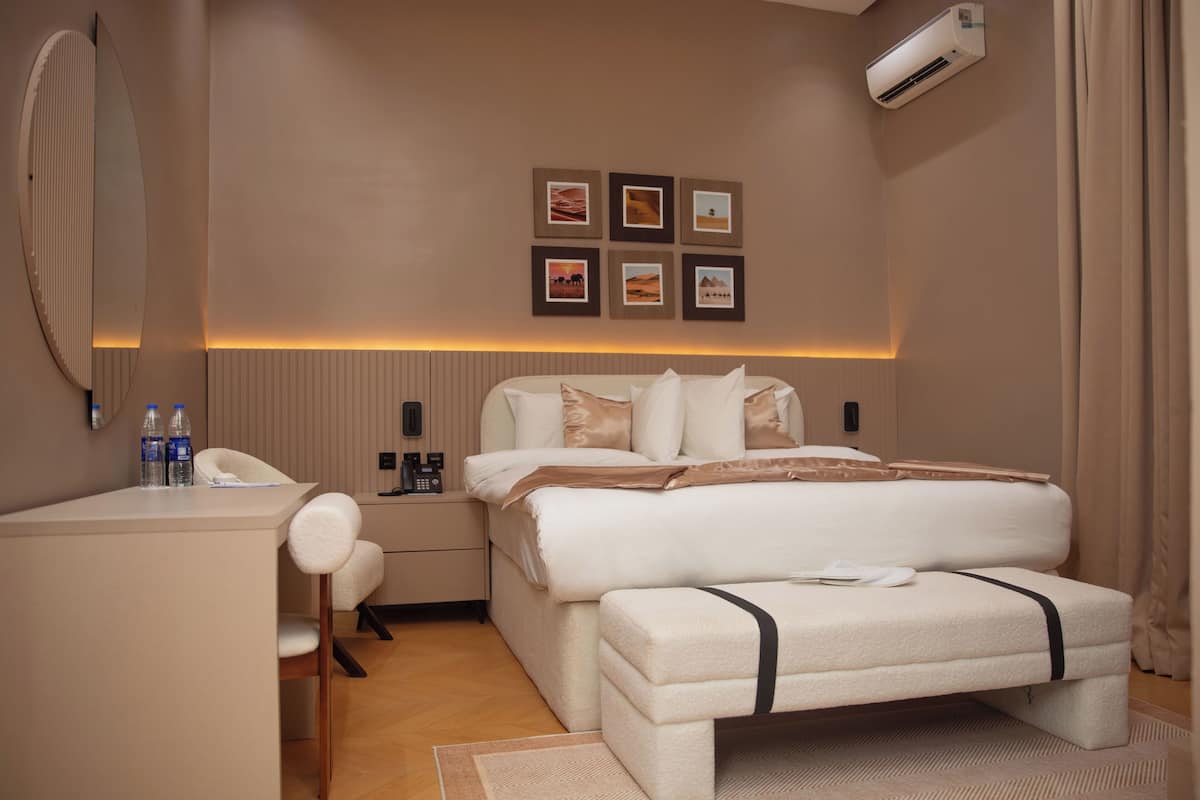 Quarto de hotel com cama, mesa de cabeceira, cadeira e escrivaninha. Há quadros nas paredes, ar condicionado e uma janela com cortinas.