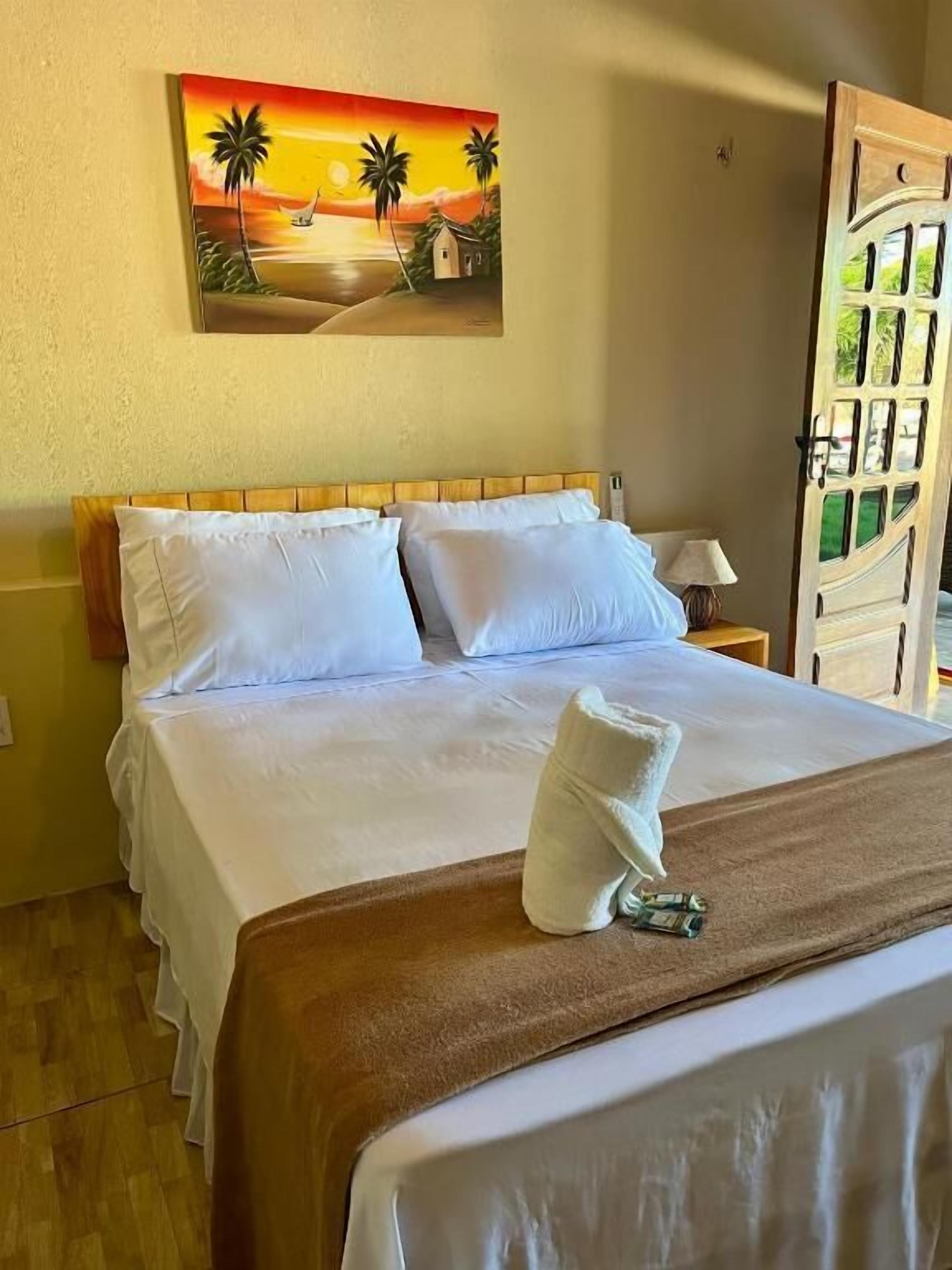 Quarto de hotel com uma cama, uma pintura de pôr do sol, uma porta de madeira e uma mesinha de cabeceira com uma luminária.