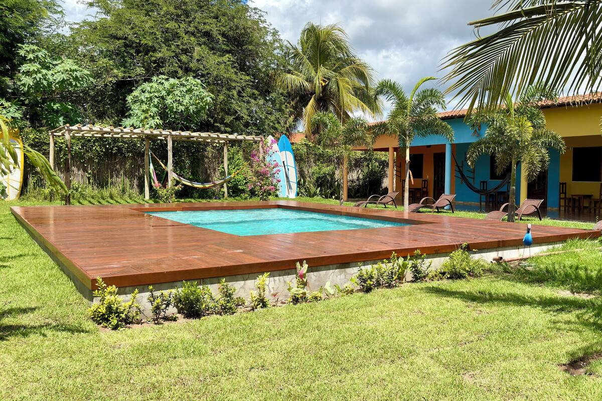 Uma piscina com deck de madeira, rodeada por grama e palmeiras, com espreguiçadeiras e um prédio ao fundo.