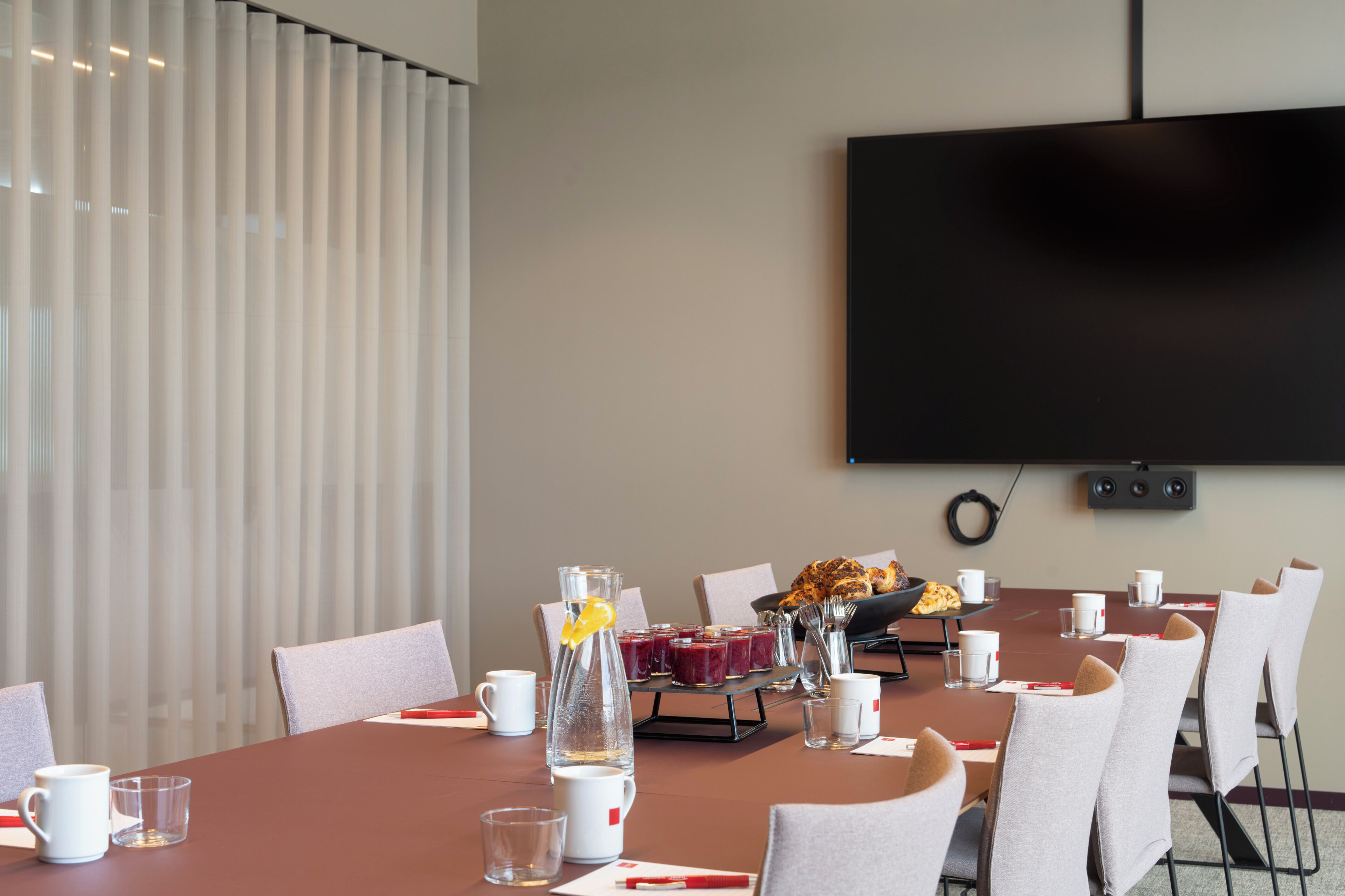 Uma sala de conferências com uma mesa grande, cadeiras, uma tela de televisão e diversos objetos sobre a mesa.