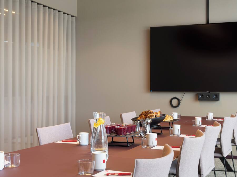 Uma sala de conferências com uma mesa grande, cadeiras, uma tela de televisão e diversos objetos sobre a mesa.