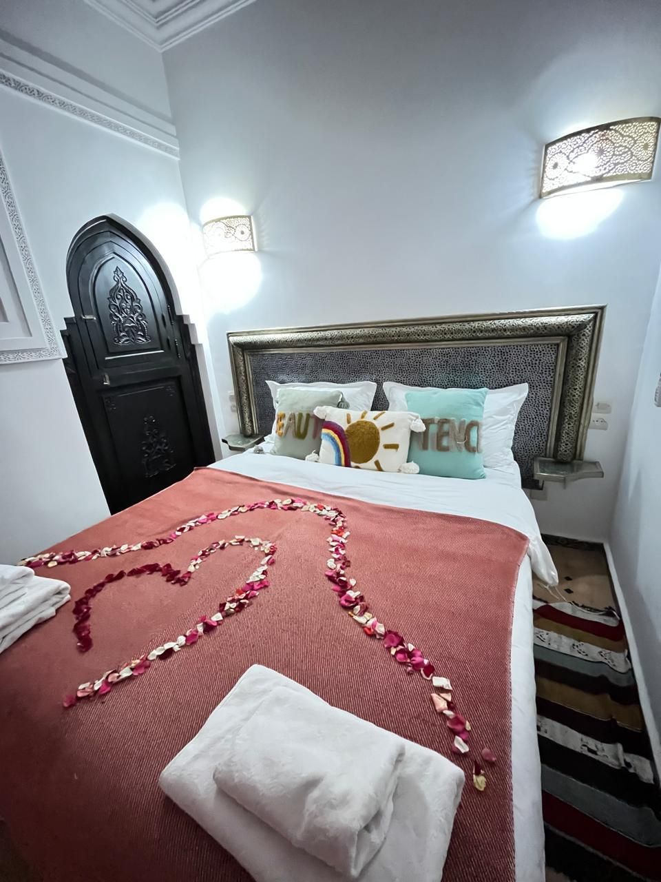 Quarto de hotel com cama, almofadas decorativas, cabeceira e luminária fixada na parede.