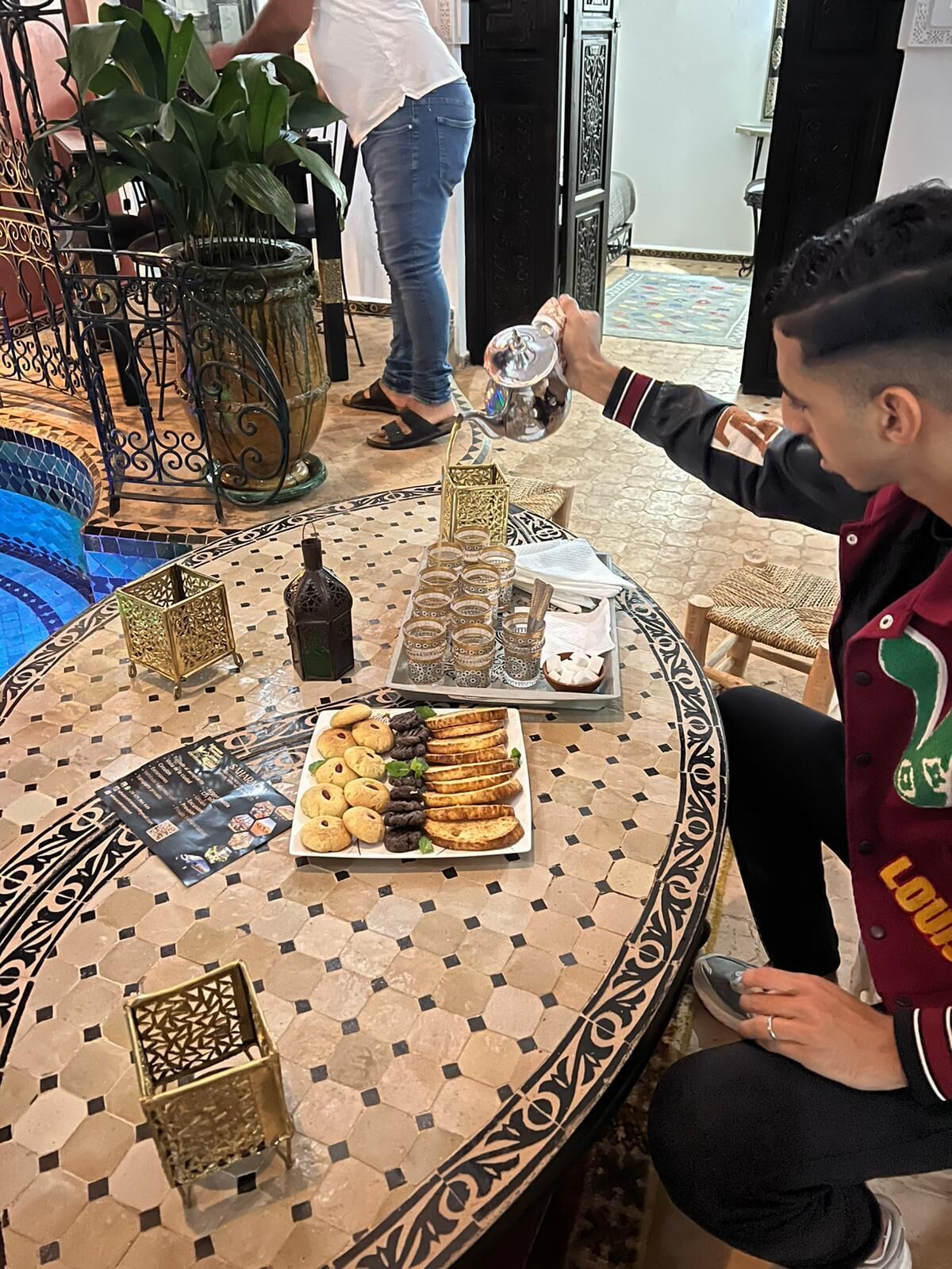 Uma mesa redonda com chão de azulejos, uma bandeja de petiscos e um bule de chá.