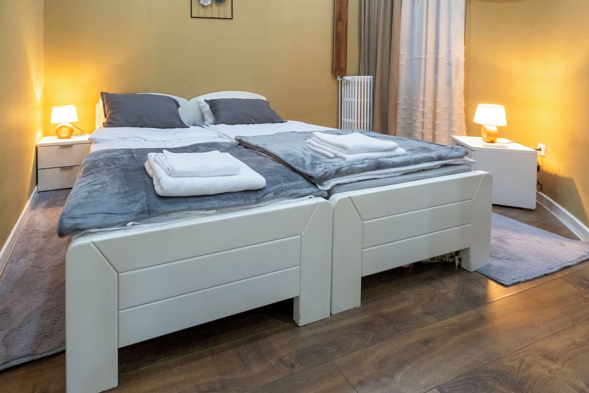 Uma cama de casal com colcha cinza e branca, duas mesas de cabeceira com luminárias, um radiador e uma janela com cortinas.