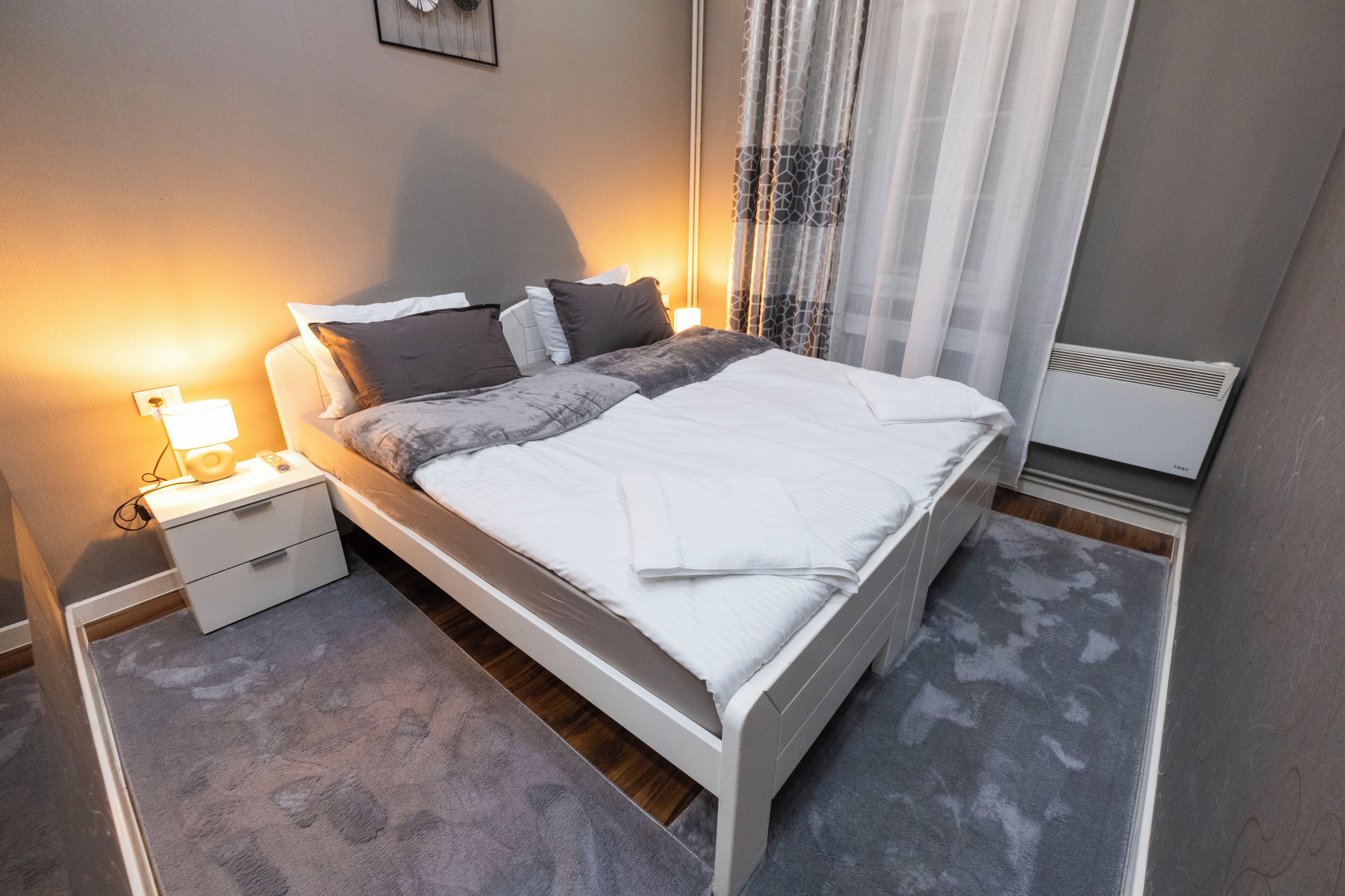 Cama bem arrumada com roupa de cama branca, mesa de cabeceira com abajur e janela com cortinas leves.