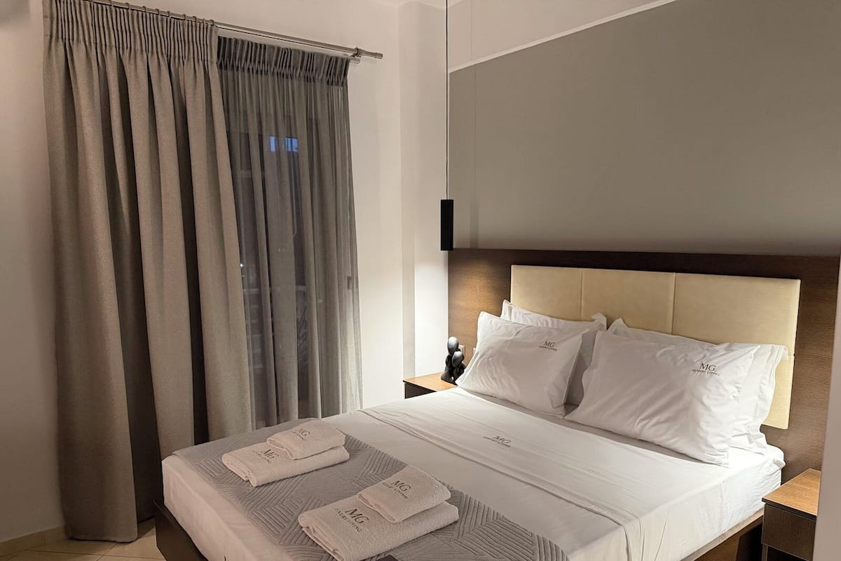 Quarto de hotel com cama, cortinas, cabeceira e mesa de cabeceira.