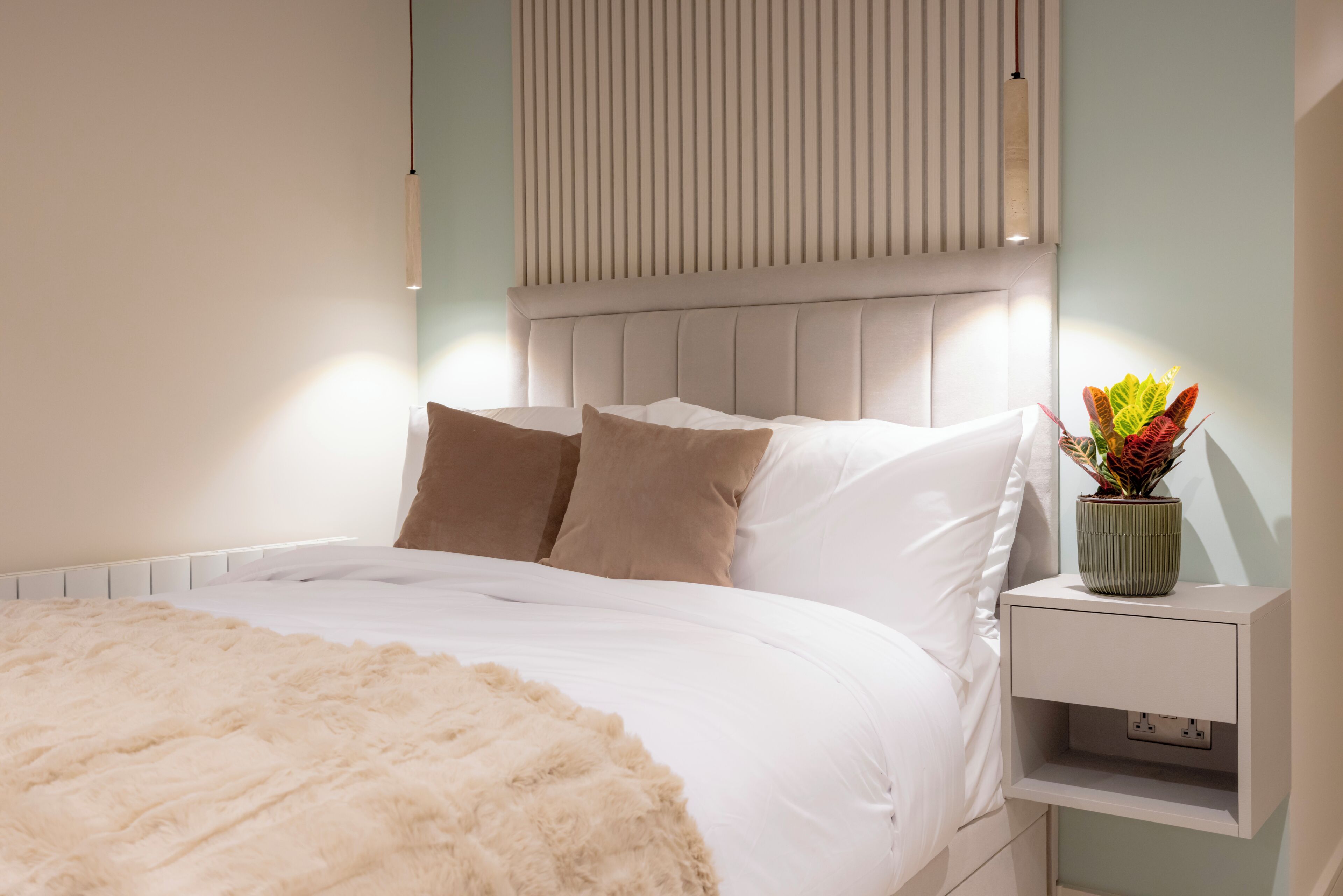 Cama bem arrumada com roupa de cama branca e um cobertor bege macio, mesa de cabeceira com uma planta em vaso, e luminária fixada na parede.