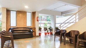 Uma área de recepção com um sofá de couro preto, algumas cadeiras de plástico, plantas em vasos e uma escada.