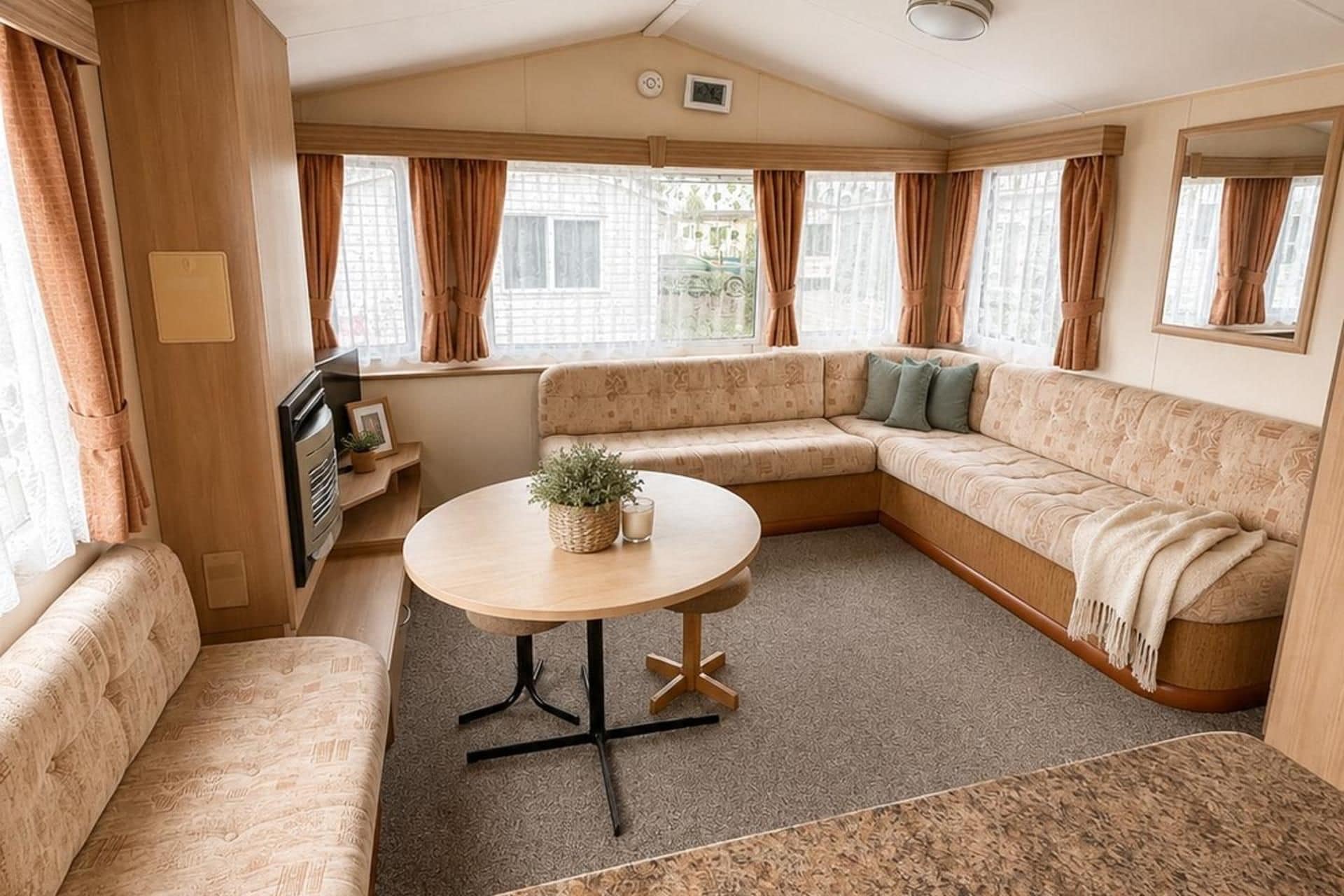 Interior acolhedor de um veículo de recreação, com sofá em formato de L, uma mesa pequena e janela com cortinas.