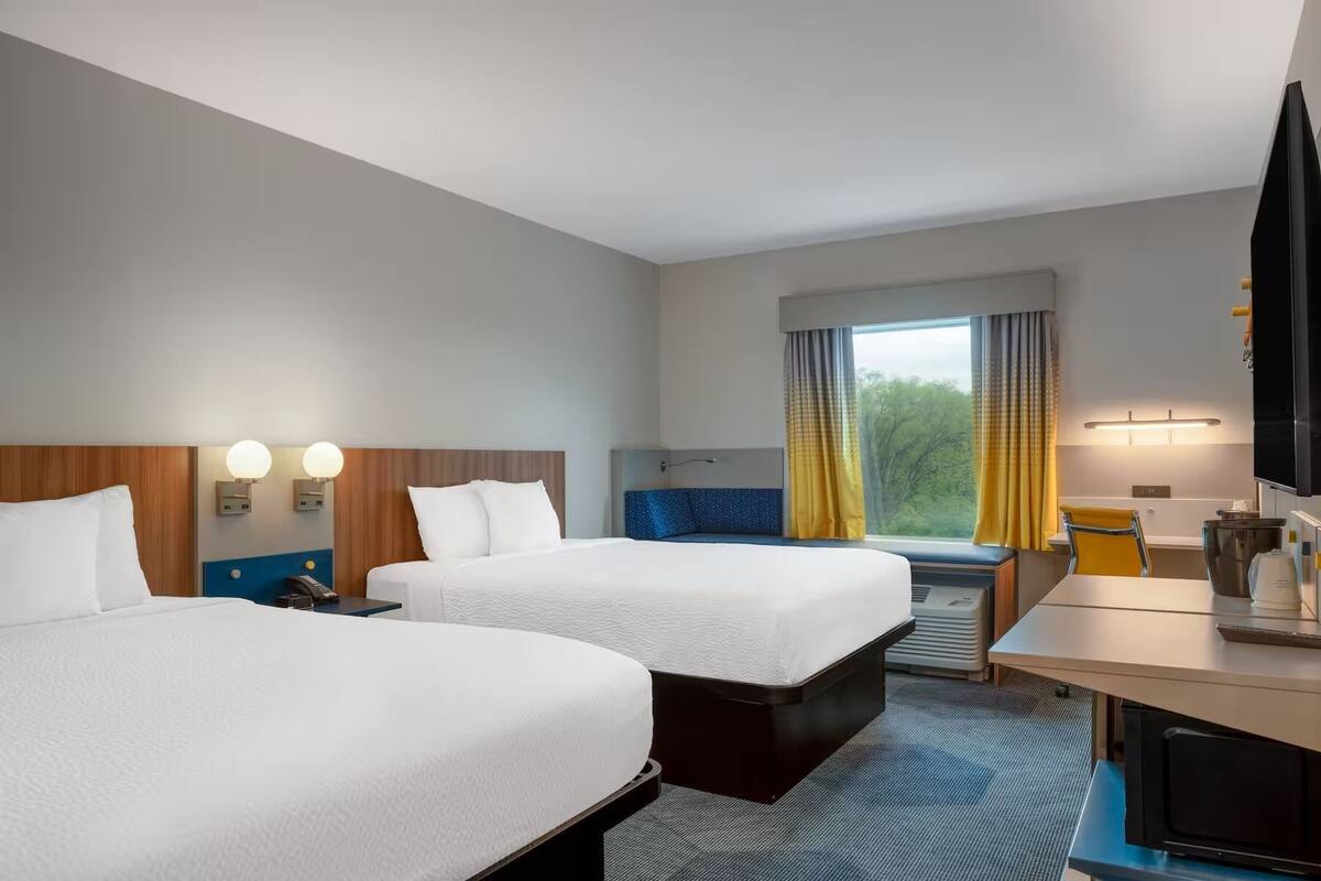 Quarto de hotel com duas camas, uma escrivaninha, televisão e janela com cortinas.