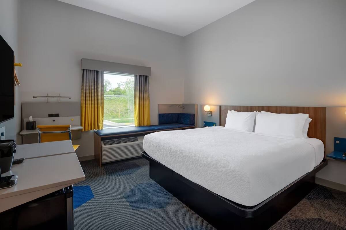 Quarto de hotel com uma cama grande, uma escrivaninha com cadeira, uma janela com cortinas e uma televisão fixada na parede.