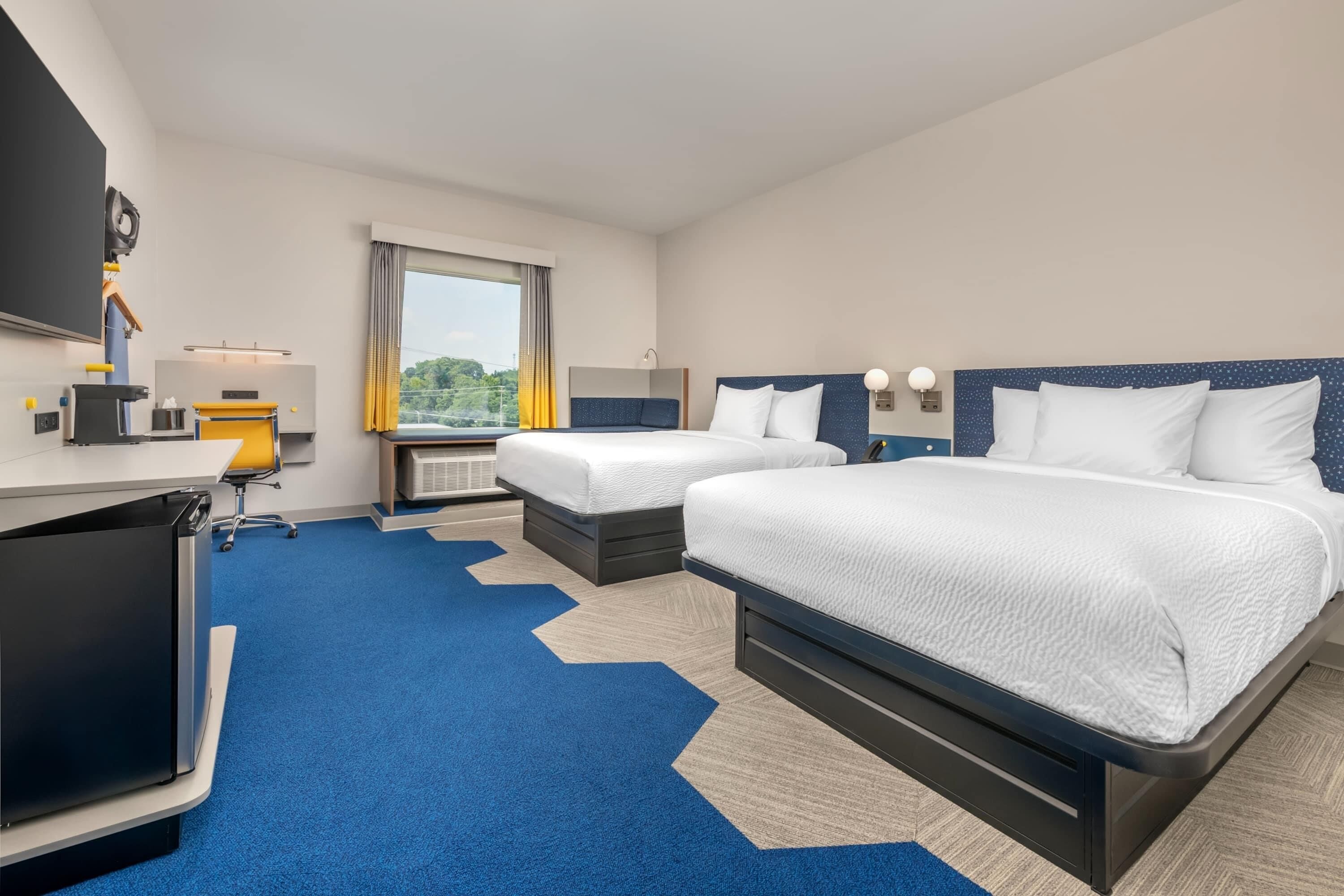Quarto de hotel com duas camas, uma escrivaninha, uma cadeira, uma televisão e uma janela com cortinas.