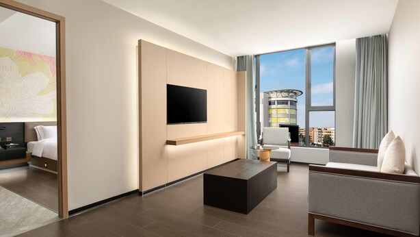Quarto de hotel moderno com televisão de tela plana, sofá, mesa de centro e vista para a cidade através de grandes janelas.