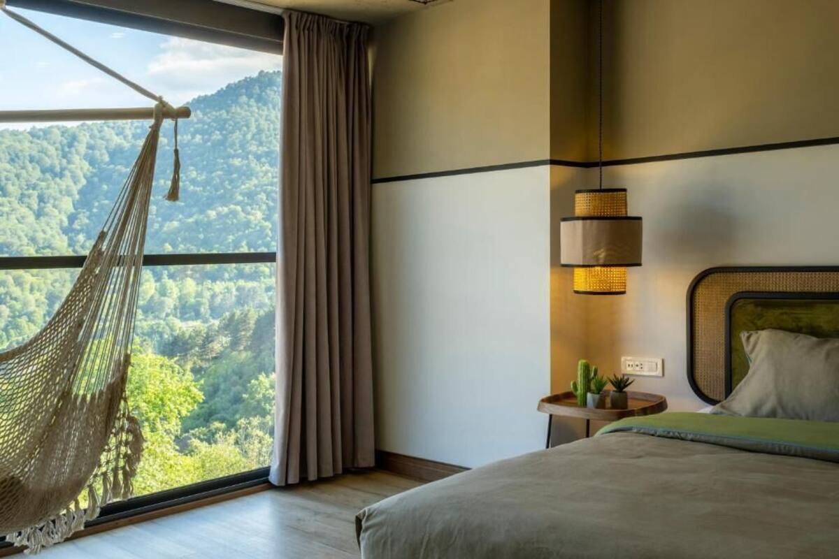 Quarto com cama, mesa de cabeceira, abajur, cadeira suspensa e vista para uma área arborizada.