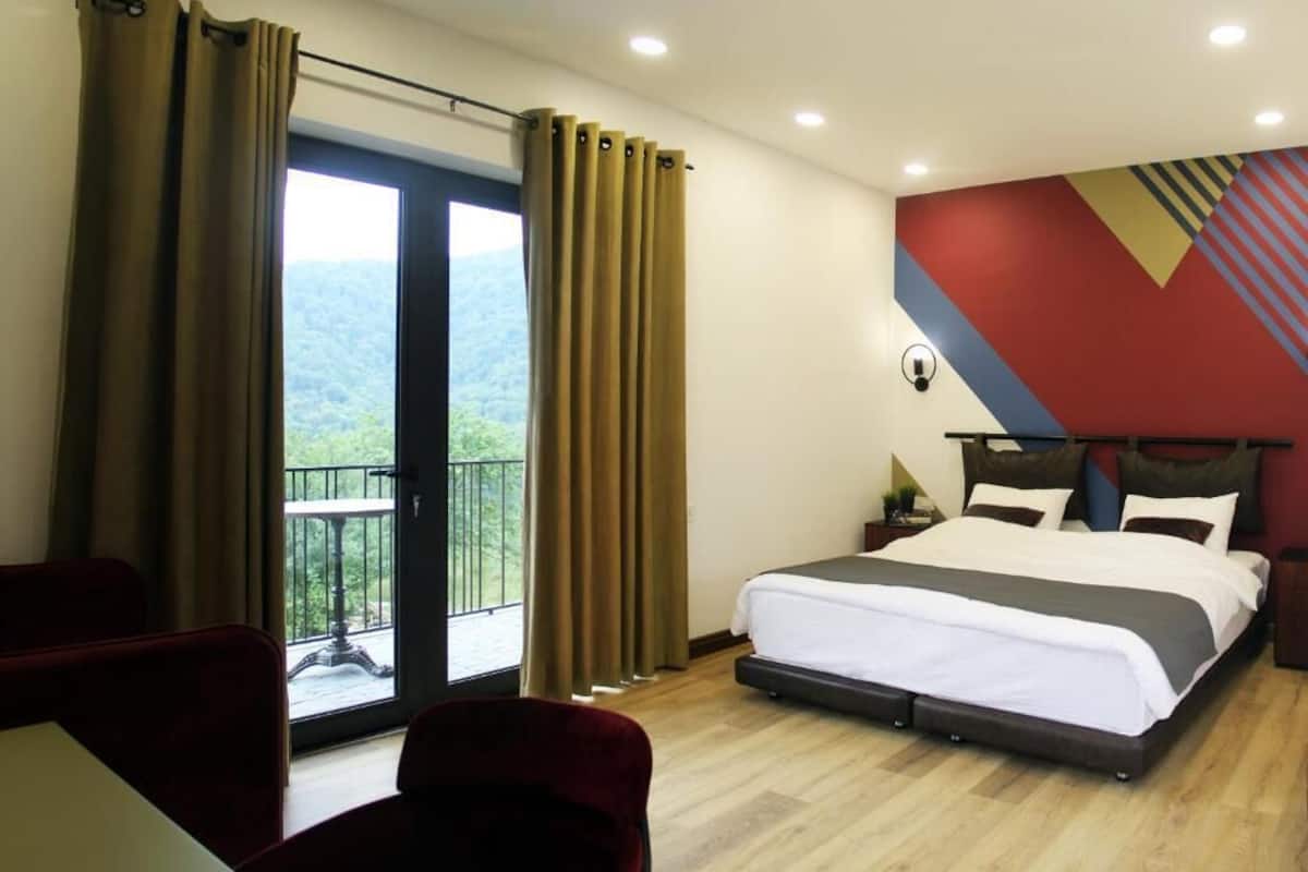 Quarto de hotel com cama, varanda com cortinas, parede vermelha e vista para o verde.