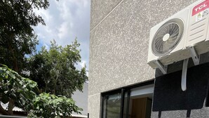 Fachada de edifício com um pátio ladrilhado, uma pequena árvore e um aparelho de ar condicionado fixado na parede.