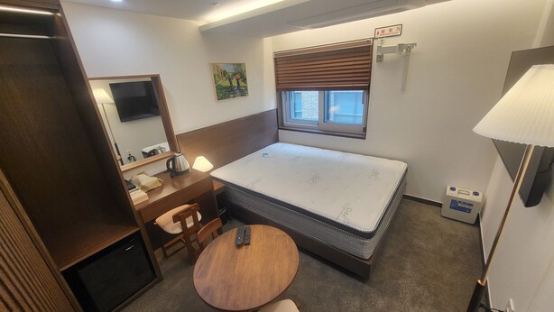 Quarto de hotel compacto com cama, escrivaninha, cadeira, mesinha, televisão, luminária e janela com persianas.