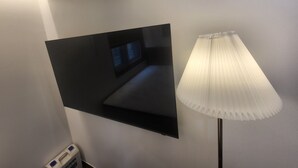 Televisão de tela plana presa na parede, um abajur de chão e uma pequena janela com persianas.