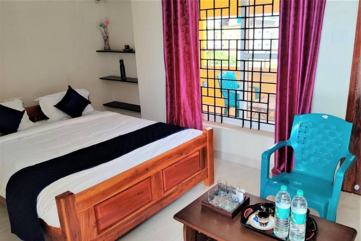 Quarto com cama de madeira, uma mesinha com garrafas de água, uma cadeira e uma janela com cortinas.