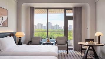 Quarto de hotel com uma cama, duas cadeiras, uma mesinha e vista para o horizonte urbano por grandes janelas.